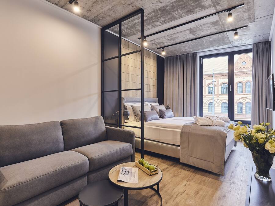 Quarto de hotel moderno com sofá cinzento, uma mesinha redonda com jornal e maçãs, cama com cabeceira cinzenta e vista para um edifício histórico através de grandes janelas.