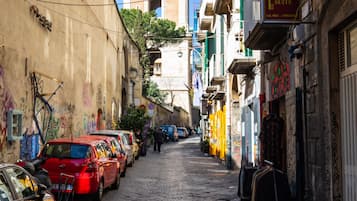 Uma rua de paralelepípedos estreita, ladeada por edifícios, com uma torre de sino ao fundo e carros estacionados nas laterais.