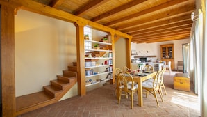 Interior em madeira com escadaria, sala de jantar com cadeiras, estante de livros e área de cozinha.