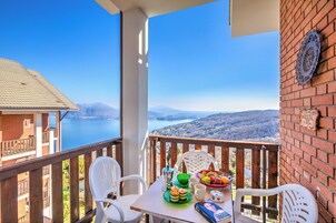Varanda com mesa posta para o café da manhã, incluindo uma tigela de frutas, uma cafeteira e um livro. A varanda oferece uma vista panorâmica de um lago e montanhas.