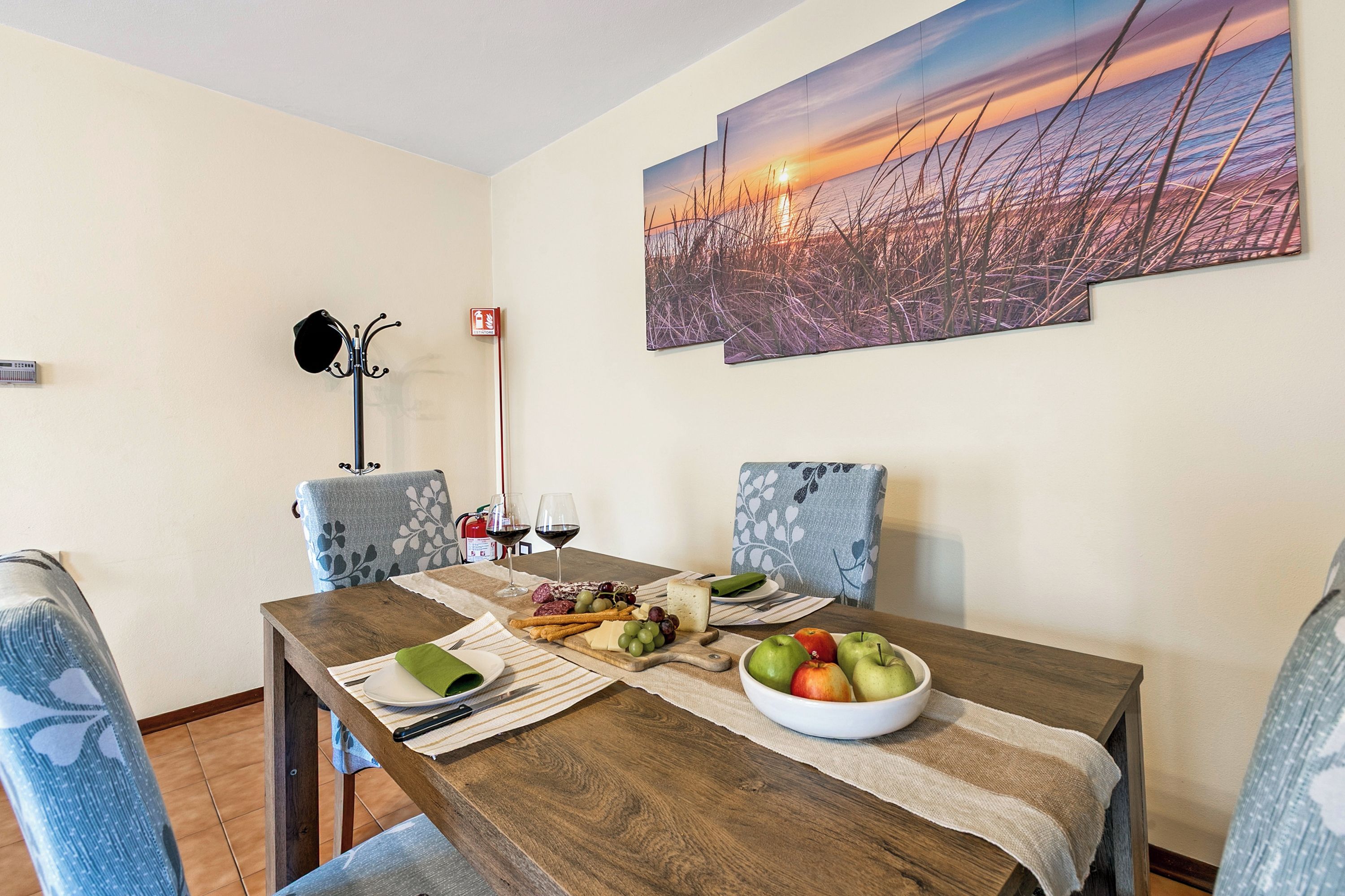 Uma sala de jantar com uma mesa de madeira posta para refeição, apresentando uma tigela de fruta, queijo e palitos de pão. Há duas cadeiras com estofamento floral e um quadro com o pôr do sol sobre a relva na parede.