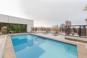 Piscina no terraço com vista desobstruída da linha do horizonte da cidade, cercada por cadeiras de descanso e fachada de um edifício moderno.