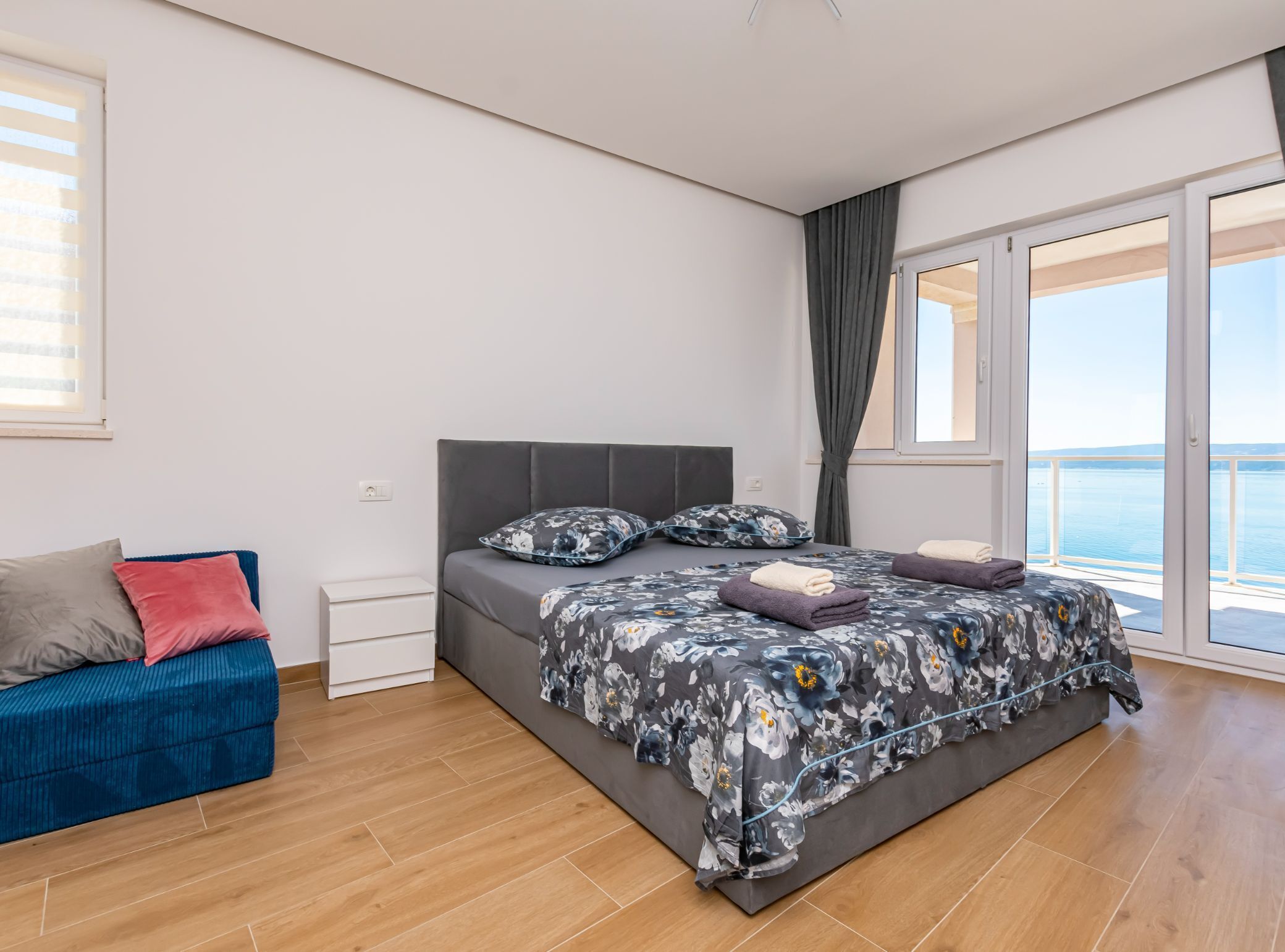 Quarto com cama, sofá azul e vista para o mar através de grandes janelas.