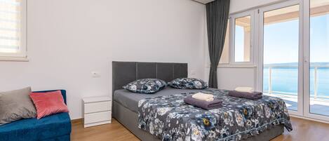 Quarto com cama, sofá azul e vista para o mar através de grandes janelas.