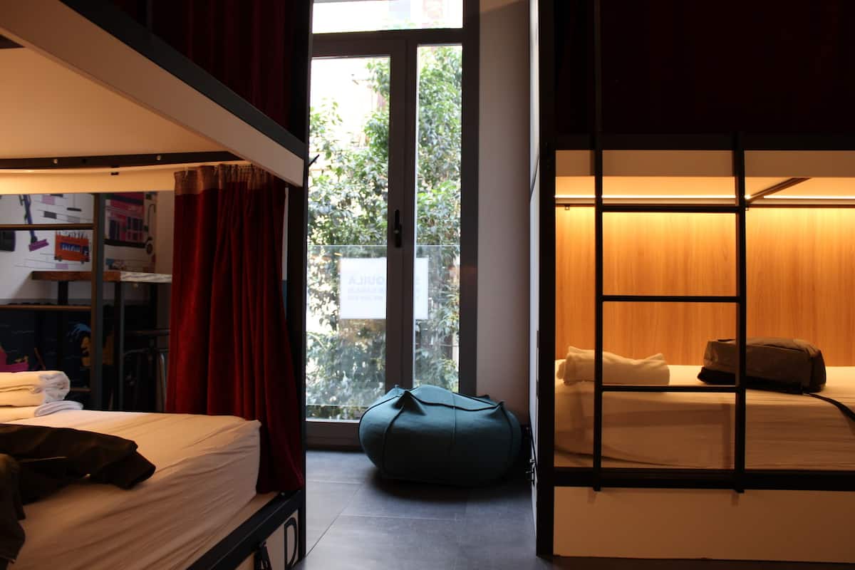 Quarto estilo dormitório com beliches, uma poltrona de encosto e uma janela com vista para vegetação.