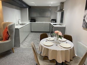 Uma cozinha moderna com armários cinza, uma mesa de jantar posta para dois com toalhas brancas e uma cadeira com almofada vermelha.