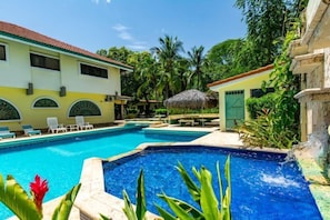 Uma piscina com espreguiçadeiras e uma estrutura com telhado de palha, cercada por vegetação exuberante e um edifício com janelas em arco.