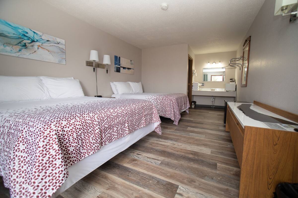 Quarto de hotel com duas camas, uma cômoda de madeira, um espelho e um banheiro visível ao fundo.