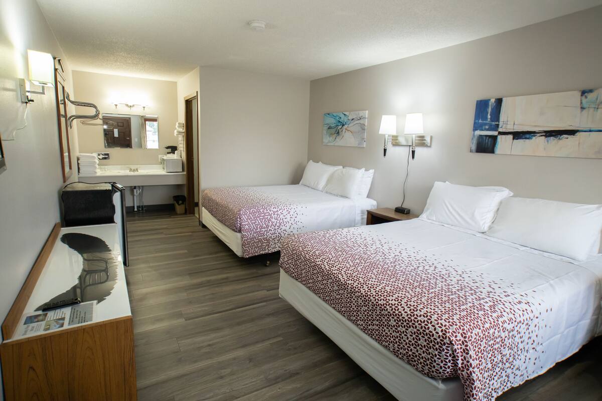 Quarto de hotel com duas camas, uma escrivaninha, um espelho e um banheiro visível ao fundo.