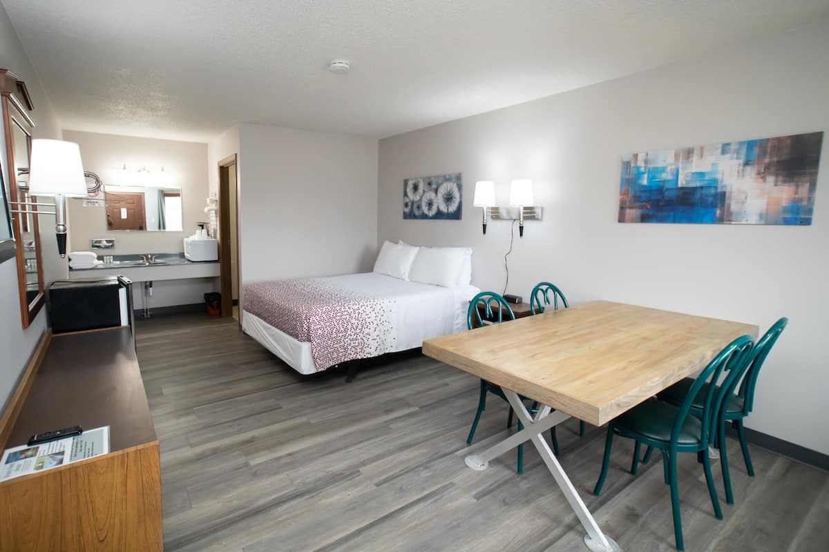 Quarto de hotel com uma cama, uma mesa de jantar de madeira com cadeiras, uma escrivaninha com um computador e um banheiro visível ao fundo.