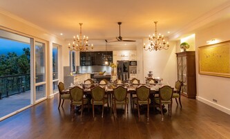 Uma sala de jantar espaçosa com uma grande mesa posta para seis pessoas, rodeada por cadeiras estofadas verdes. Uma cozinha moderna com eletrodomésticos de aço inoxidável é visível ao fundo.