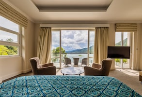 Quarto de hotel com uma cama grande, televisão, varanda com vista para o mar e montanhas, e duas cadeiras.