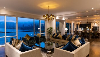 Ampla sala de estar com uma grande janela com vista para a água, um candelabro e uma área de jantar com várias cadeiras e mesas.