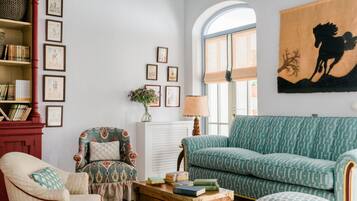 Uma sala de estar com um sofá estampado, poltronas, uma mesa de centro de madeira com livros, uma estante vermelha e quadros nas paredes.