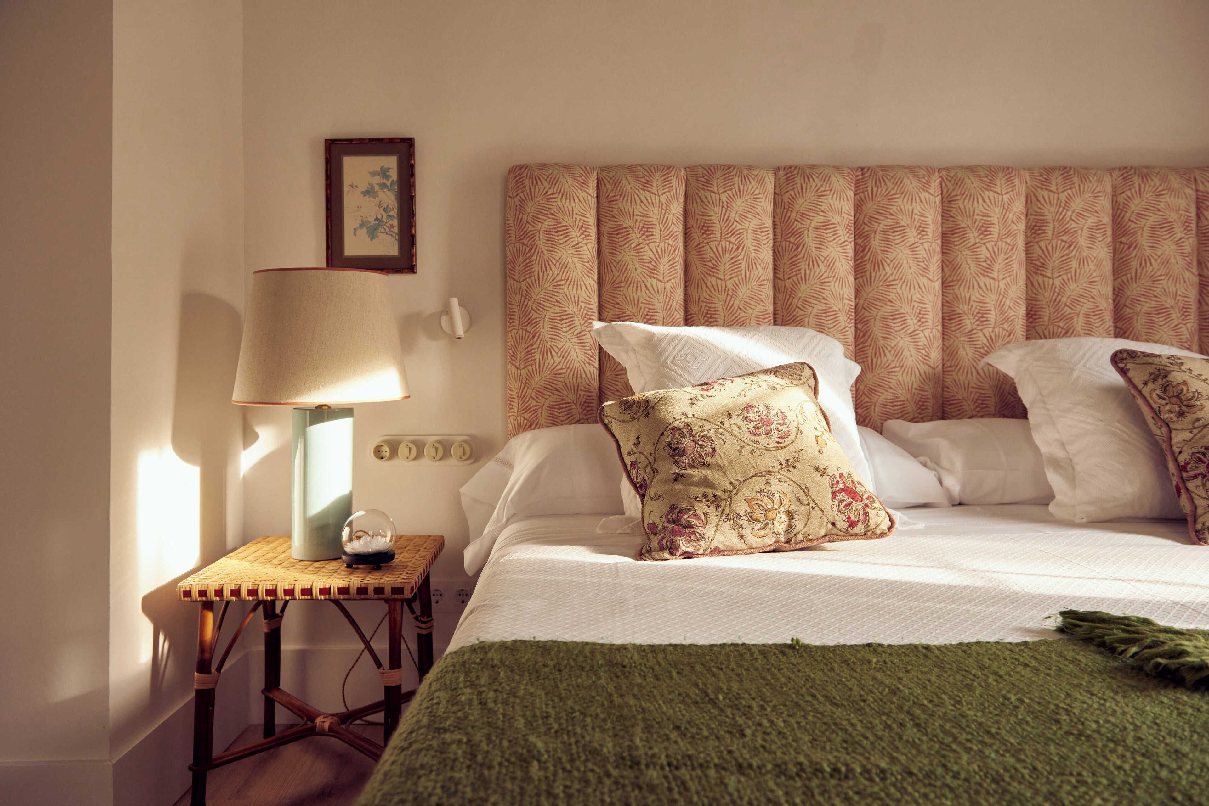 Uma cama bem arrumada com cabeceira estampada, uma luminária de mesa e uma pequena mesinha com abajur.