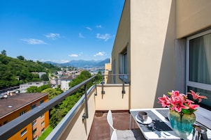 Uma varanda com vista para a paisagem urbana, uma mesa com um vaso de flores e uma janela com cortinas.