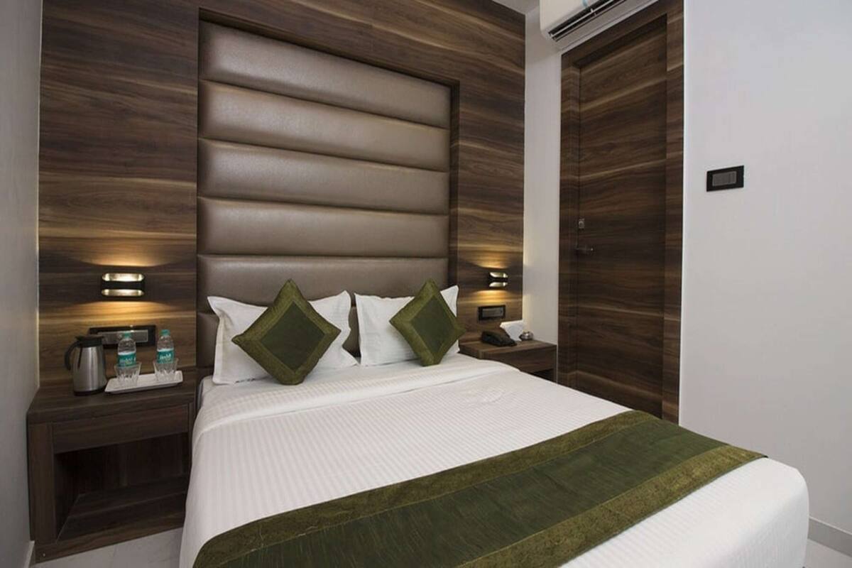 Quarto de hotel com uma cama grande, cabeceira de madeira, abajures e mesa de cabeceira com chaleira e garrafas de água.