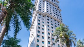 Um hotel alto com design moderno, apresentando um pátio central com uma fonte, palmeiras e uma placa indicando comodidades como restaurante, salas de reunião e academia.