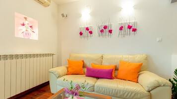 Uma sala de estar com sofá bege, almofadas laranja e rosa, mesa de centro de vidro com um vaso e decoração na parede.