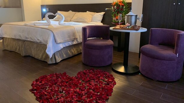 Quarto de hotel com uma cama, duas poltronas roxas, uma mesa redonda pequena com um arranjo de flores e um tapete em formato de coração feito de pétalas vermelhas no chão.