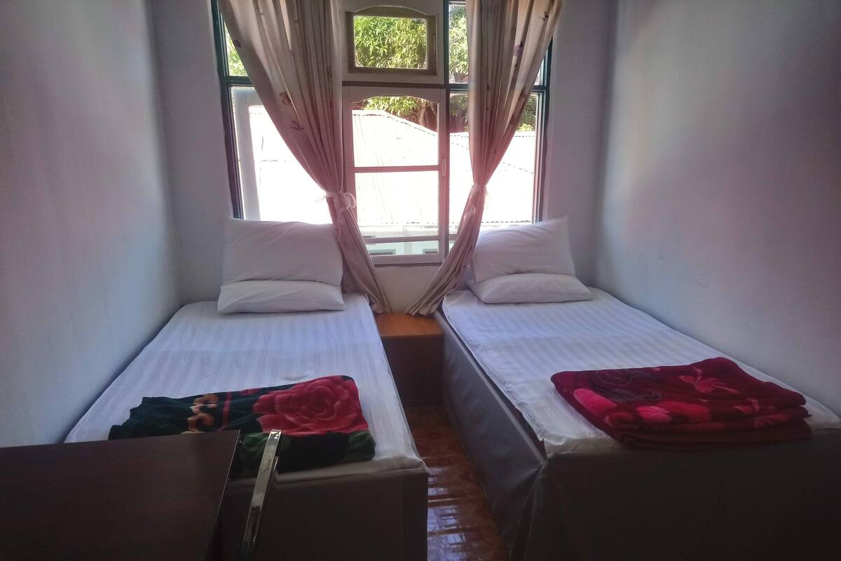 Quarto pequeno com duas camas de solteiro, uma janela com cortinas e piso de madeira.