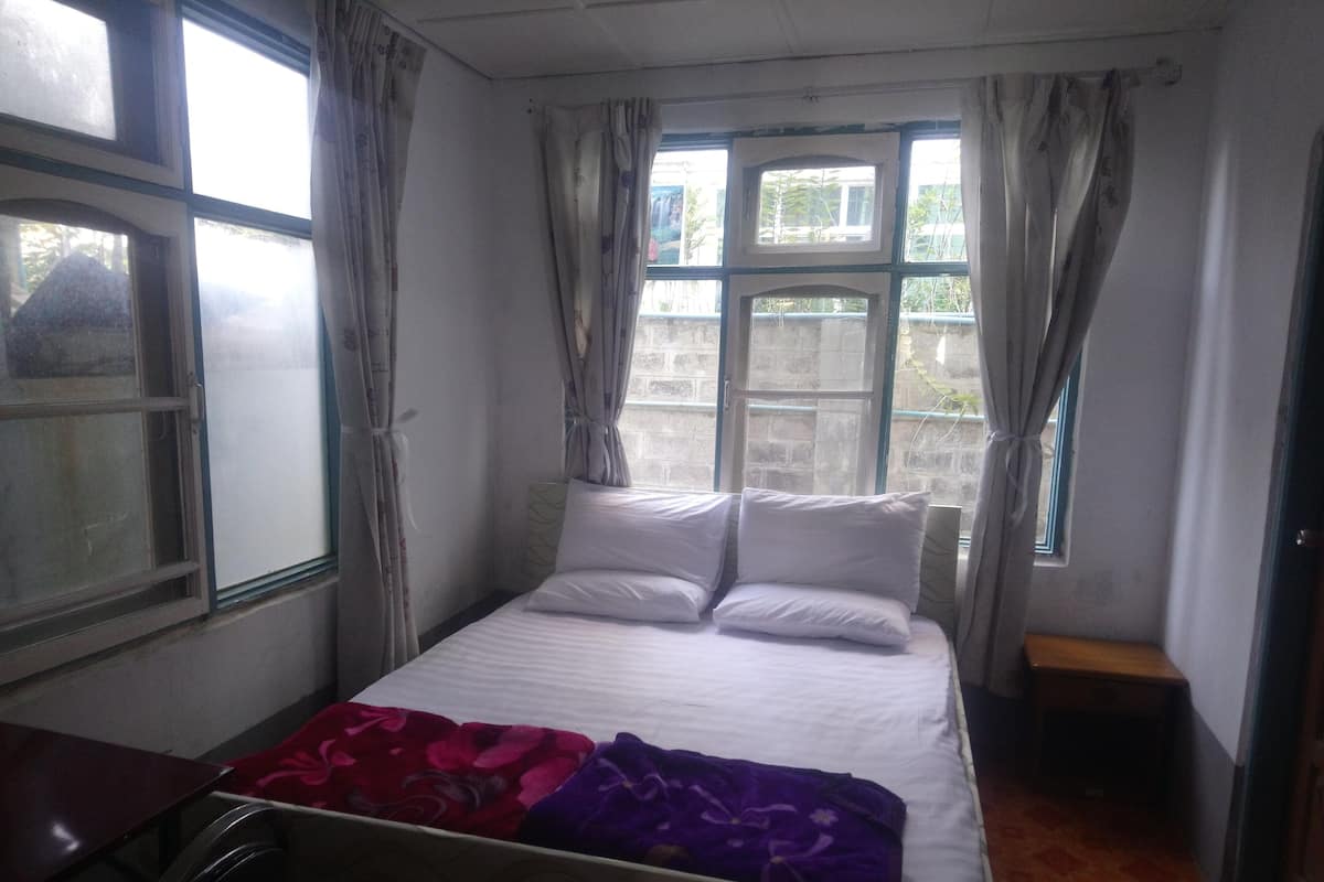 Uma cama de solteiro com roupa de cama branca num quarto com janela, cortinas e uma pequena mesa de cabeceira.
