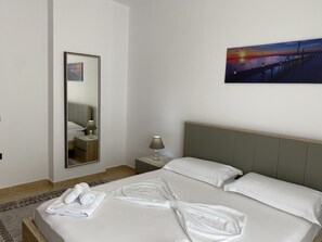 Uma cama bem arrumada com lençóis e toalhas brancas, um abajur, um espelho e uma pintura de um pôr do sol sobre um cais.