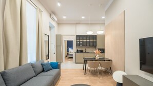 Uma sala de estar moderna com um sofá cinzento, um espaço de refeições com mesa e cadeiras, e uma cozinha ao fundo.