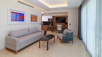 Uma sala moderna com sofá, poltrona e mesa de centro. Há uma área de cozinha com armários e uma televisão de tela plana.