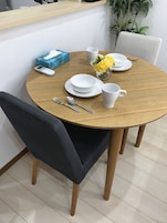 Uma mesa de jantar redonda de madeira com pratos brancos, uma caneca branca, uma colher e um garfo. Há uma caixa azul com um desenho floral e um pequeno vaso com flores amarelas.