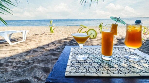 Cenário de praia com uma mesa preparada para bebidas, uma cadeira branca e vista para o oceano.
