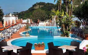 Uma área de piscina com espreguiçadeiras e guarda-sóis, cercada por vegetação e colinas ao fundo.