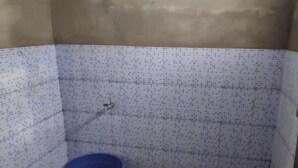 Um banheiro pequeno com azulejos estampados em azul e branco, um chuveiro e um balde azul.