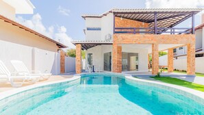 Uma piscina com espreguiçadeiras, um alpendre coberto e um edifício de dois andares com telhado de azulejos.