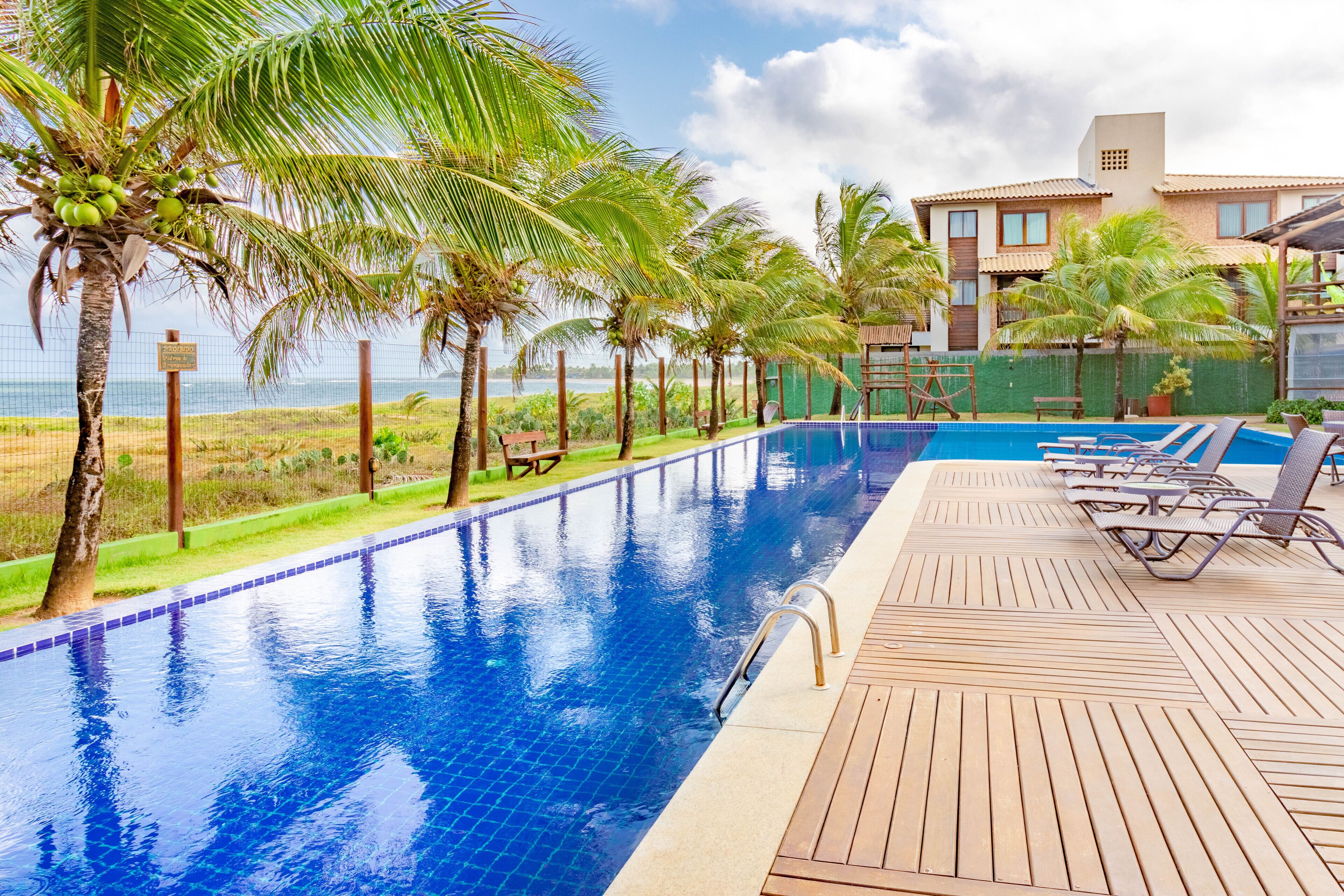 Piscina com deck de madeira e espreguiçadeiras, cercada por palmeiras e vista para o mar.