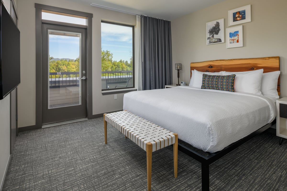 Quarto de hotel com uma cama grande, um banco, uma televisão, janela com vista e obras de arte na parede.