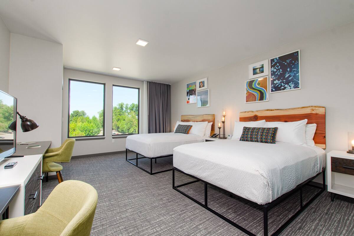 Quarto de hotel com duas camas, uma escrivaninha, uma cadeira e janelas amplas com vista para o verde.