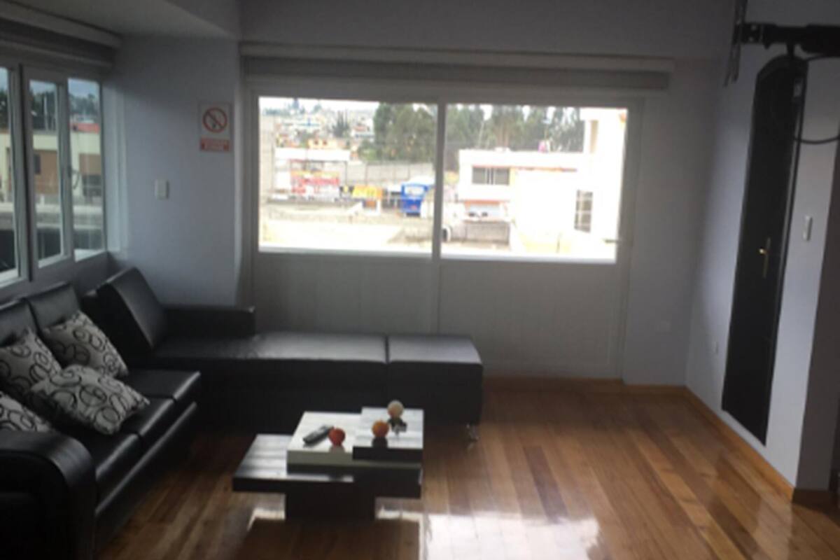 Uma sala de estar moderna com um sofá preto, uma mesa de centro e uma janela grande com vista para prédios.