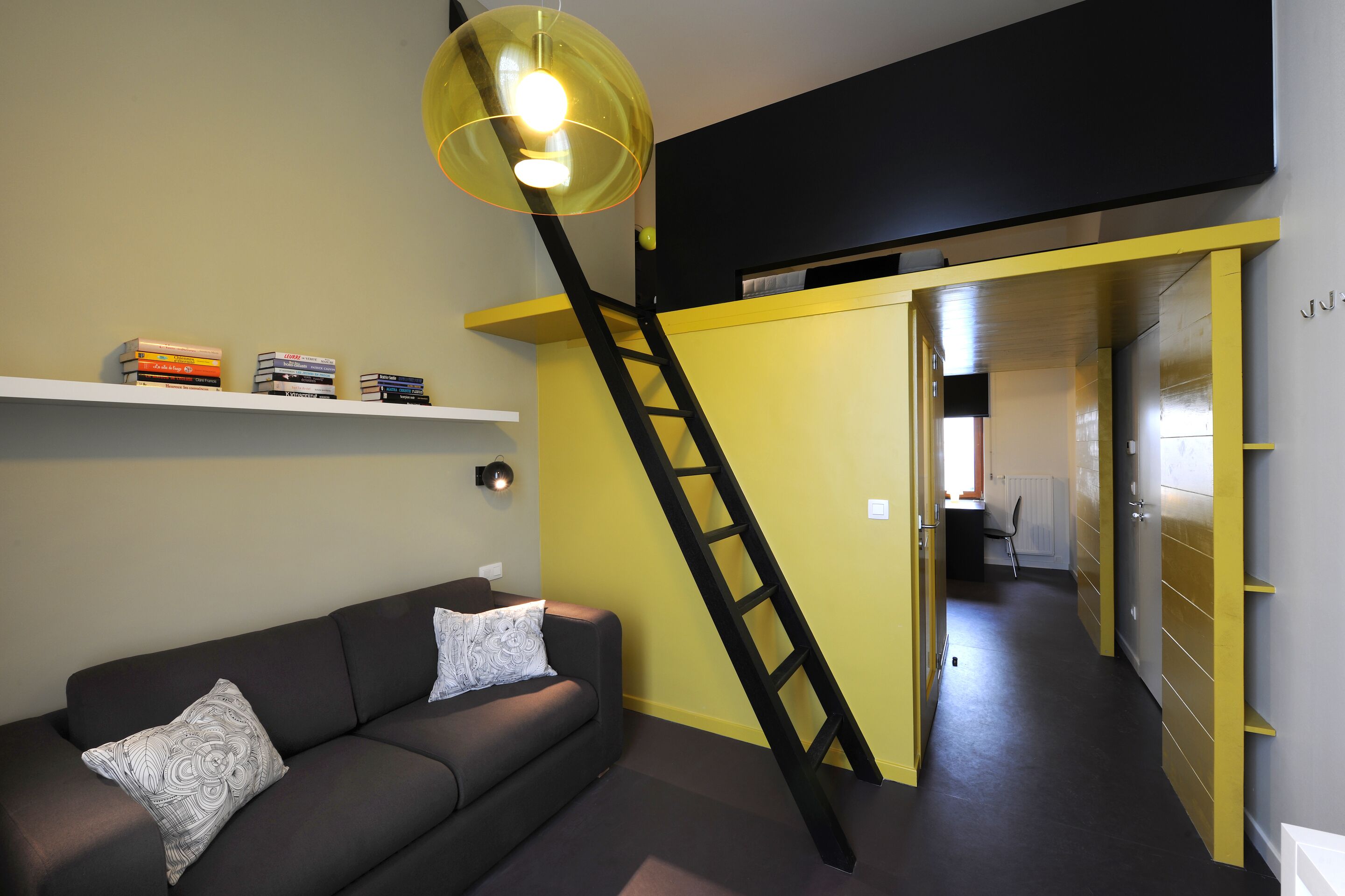 Uma sala de estar moderna com um sofá escuro, uma estante branca com livros, uma escada amarela e um lance de escadas levando a outro andar.
