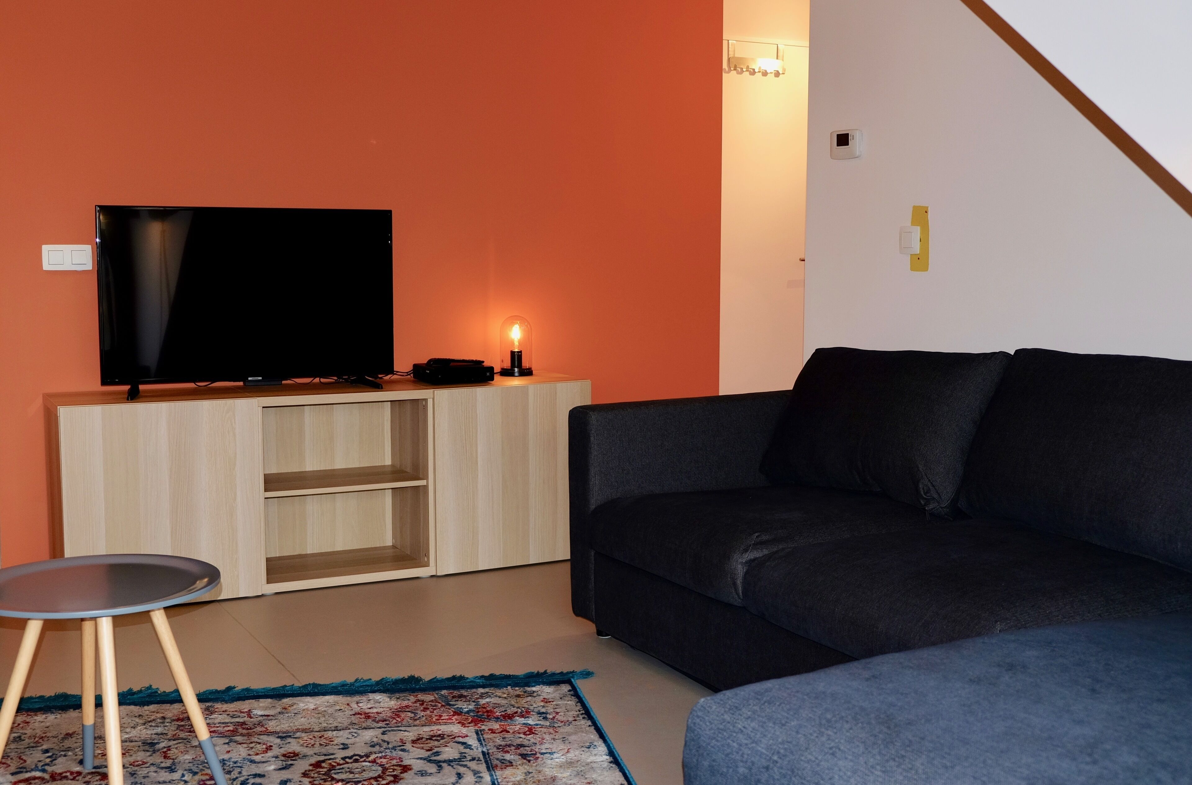 Uma sala de estar com televisão de tela plana em um aparador de madeira, um sofá cinza escuro e uma mesa redonda pequena com um tapete estampado em azul e vermelho.