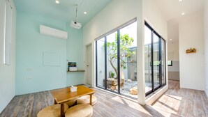 Quarto amplo com mesa de madeira e pufes trançados, grandes portas de vidro deslizantes que dão para um jardim, e projetor fixado no teto.