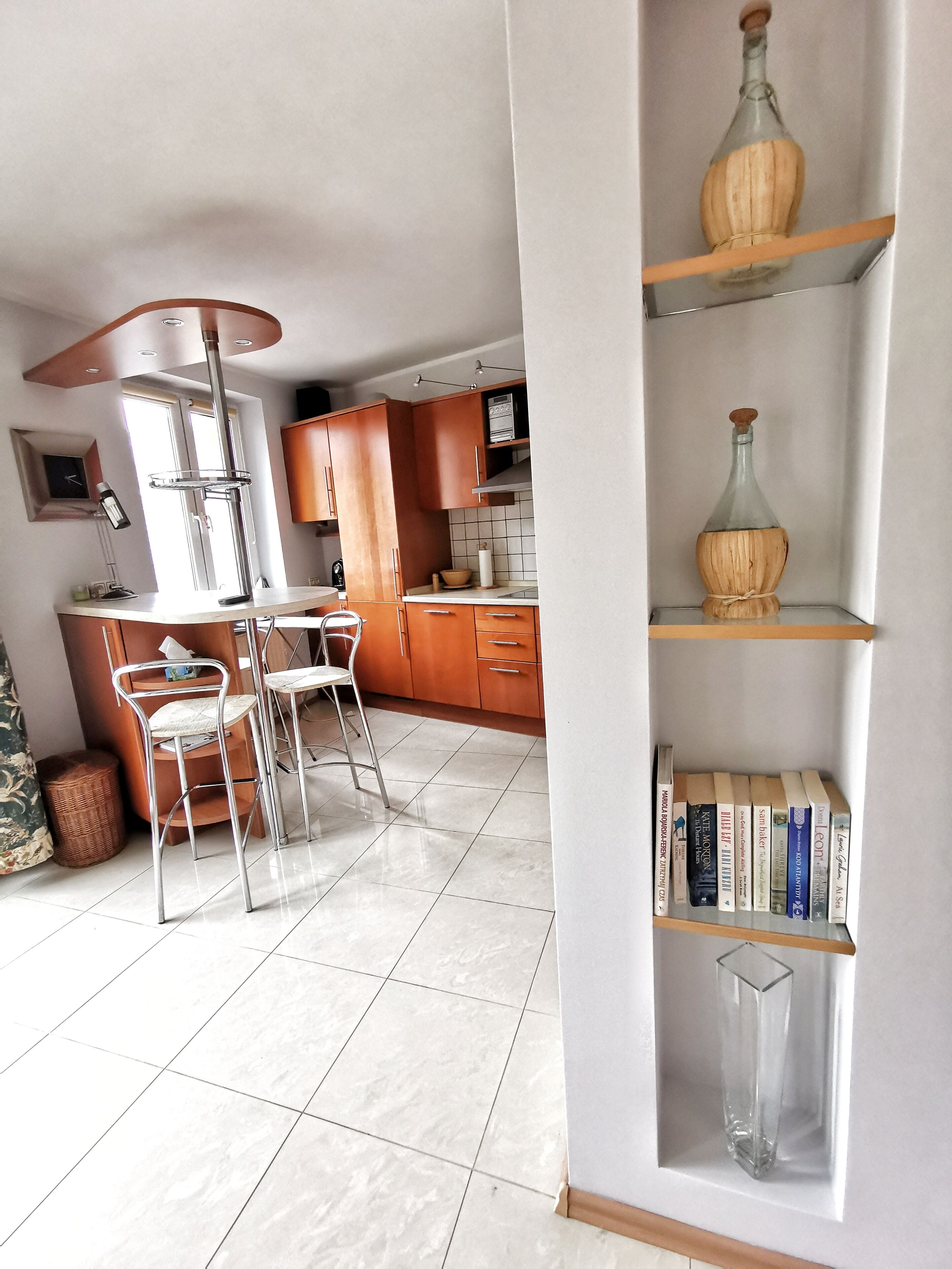 Uma cozinha moderna com armários de madeira, um espaço de refeições com cadeiras de metal e uma estante com livros e objetos decorativos.