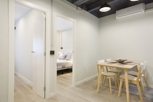 Quarto moderno e minimalista com mesa e cadeiras de madeira, cama com roupa de cama estampada e unidade de ar condicionado na parede.