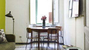 Uma sala de jantar com uma mesa redonda, quatro cadeiras, um vaso com flores, um abajur, um sofá e uma mesinha com um livro e um controle remoto.