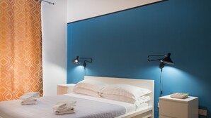 Cama bem arrumada com lençóis e almofadas brancas, duas mesas de cabeceira com abajures, e uma parede azul como detalhe.