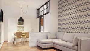 Uma sala de estar moderna com um sofá modular cinza claro, um tapete azul e uma área de jantar com cadeiras de madeira e uma mesa pequena.