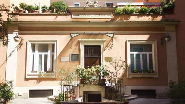 Fachada de edifício com entrada central, plantas em vasos e uma placa com a inscrição 'CASA DEI BAMBINI'.