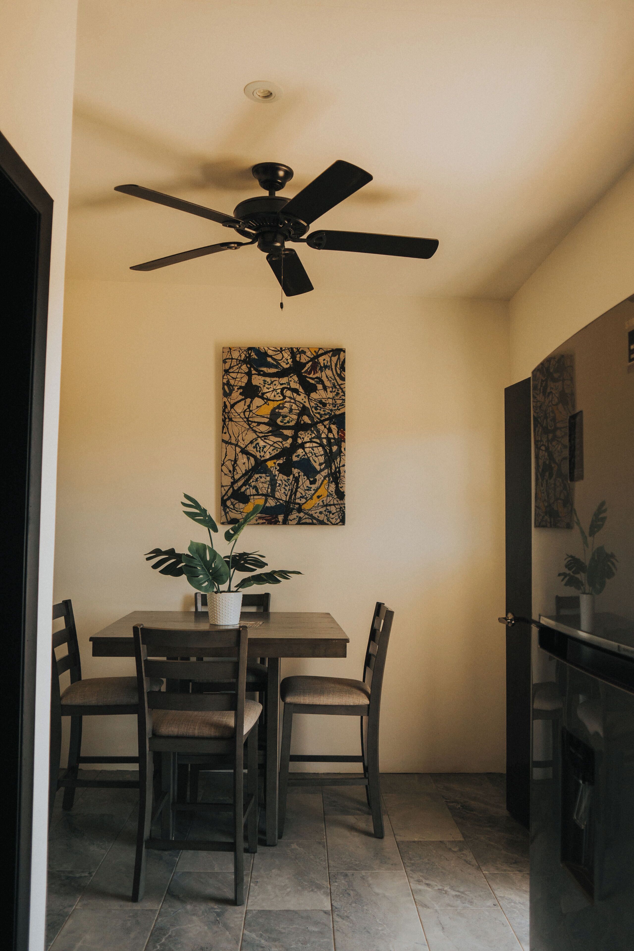Uma sala de jantar com ventilador de teto, mesa e cadeiras, planta em vaso e parede com arte abstrata.