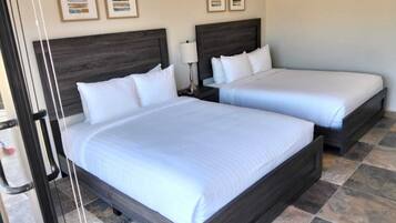 Duas camas com estrutura de madeira escura e roupa de cama branca, um criado-mudo com abajur, e uma obra de arte na parede.