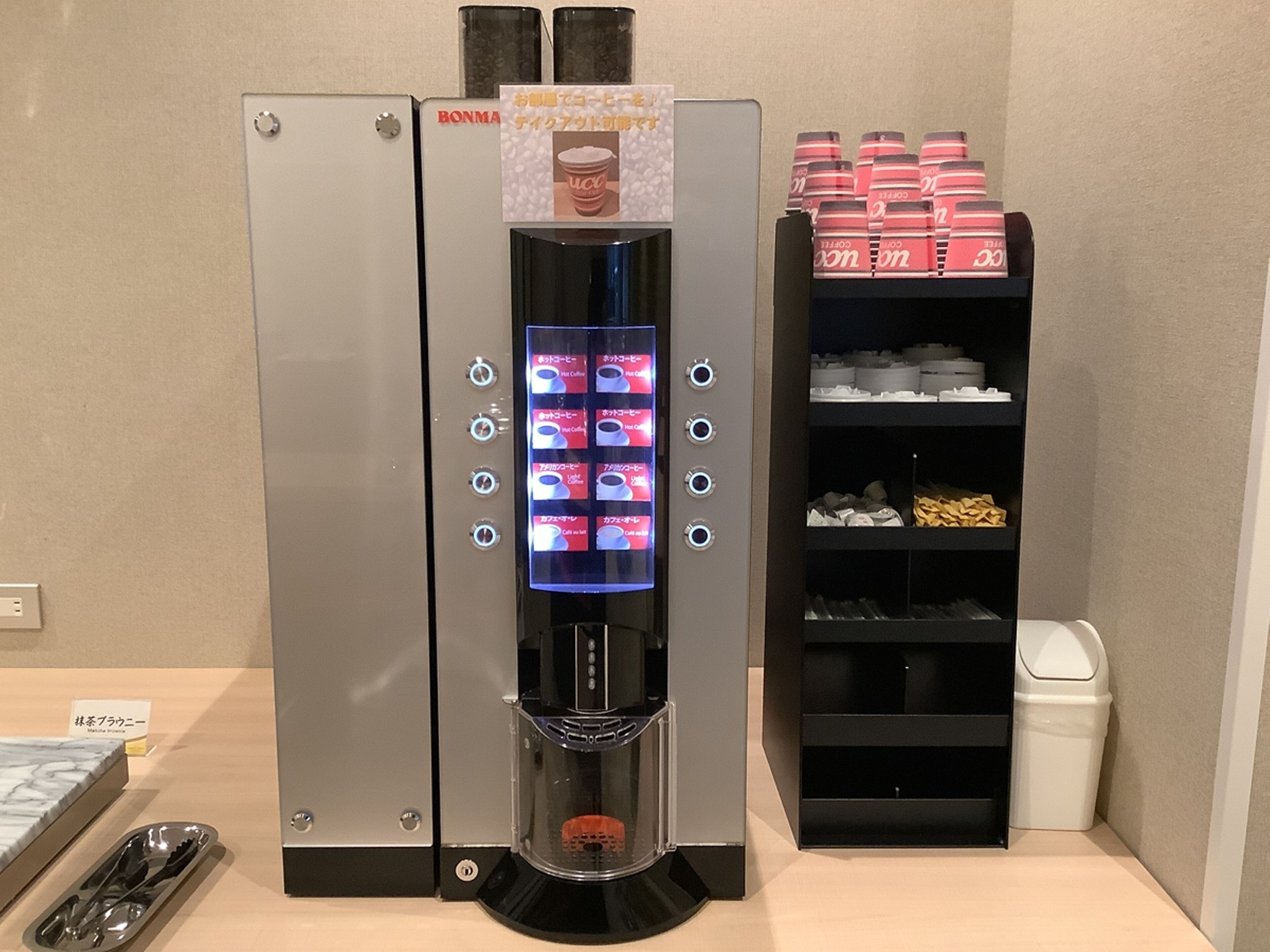 Máquina de café com visor digital e vários botões, ao lado de uma prateleira com xícaras e um cesto de lixo.